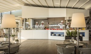 Uma cozinha moderna com chefs, um balcão repleto de pratos e uma área de refeições com cadeiras e mesas.
