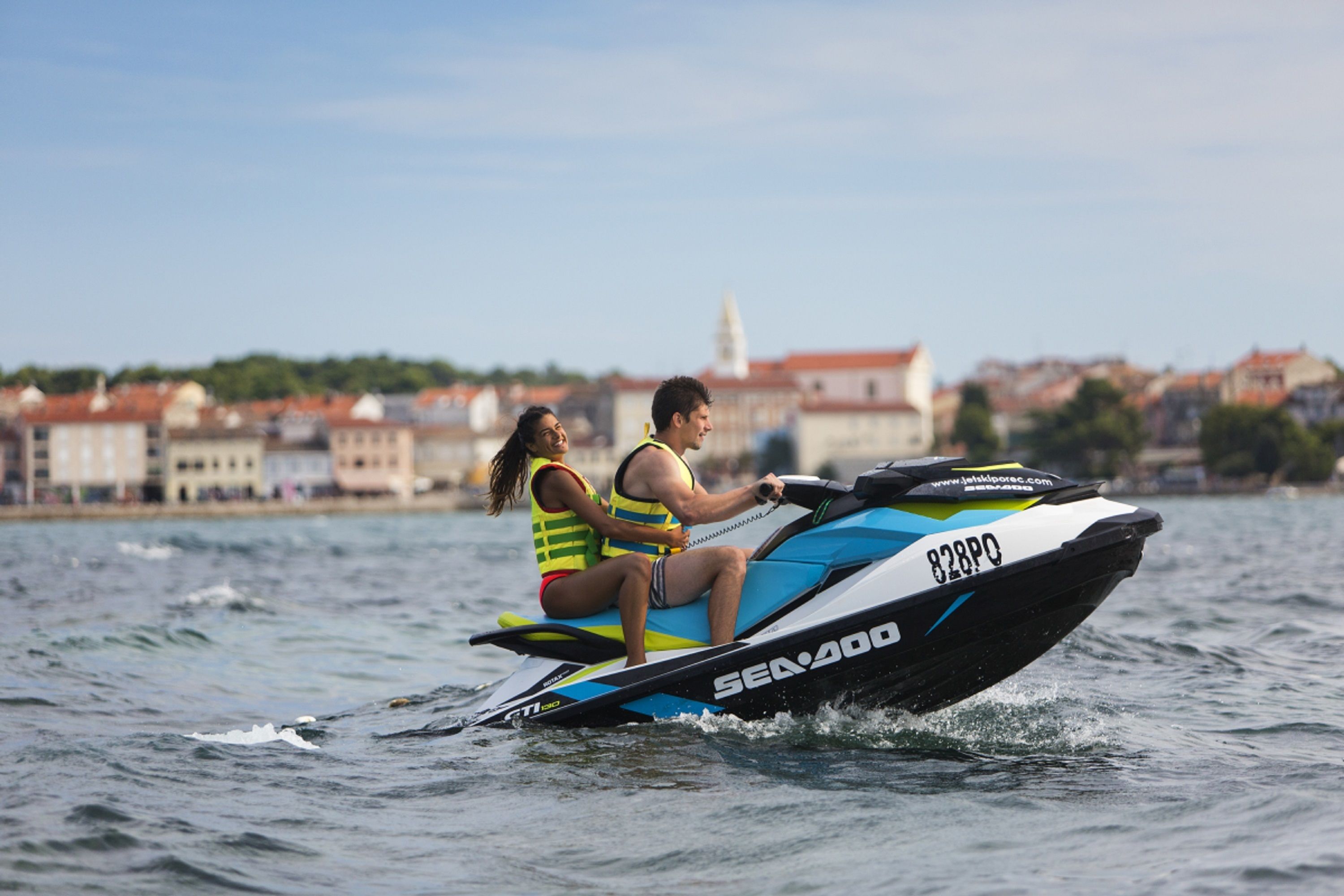 Duas pessoas em um jet ski Sea-Doo, com um horizonte urbano ao fundo.