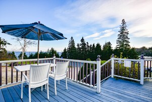 Um deck com mesa e cadeiras, um guarda-sol azul e um corrimão com vista para a água e árvores.