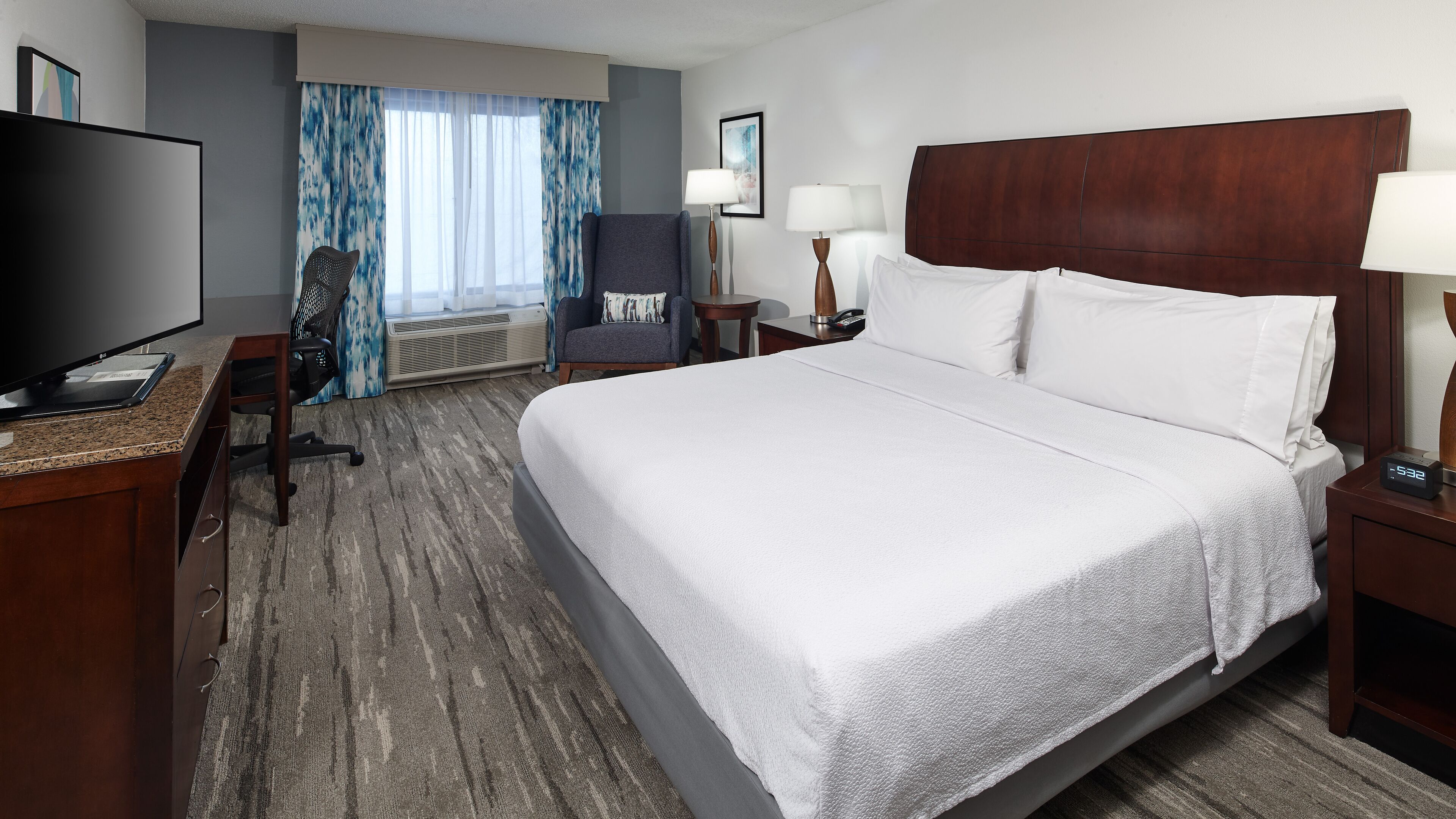 Quarto de hotel com uma cama grande, uma escrivaninha com televisão de tela plana, uma cadeira, duas luminárias de cabeceira e uma janela com cortinas.