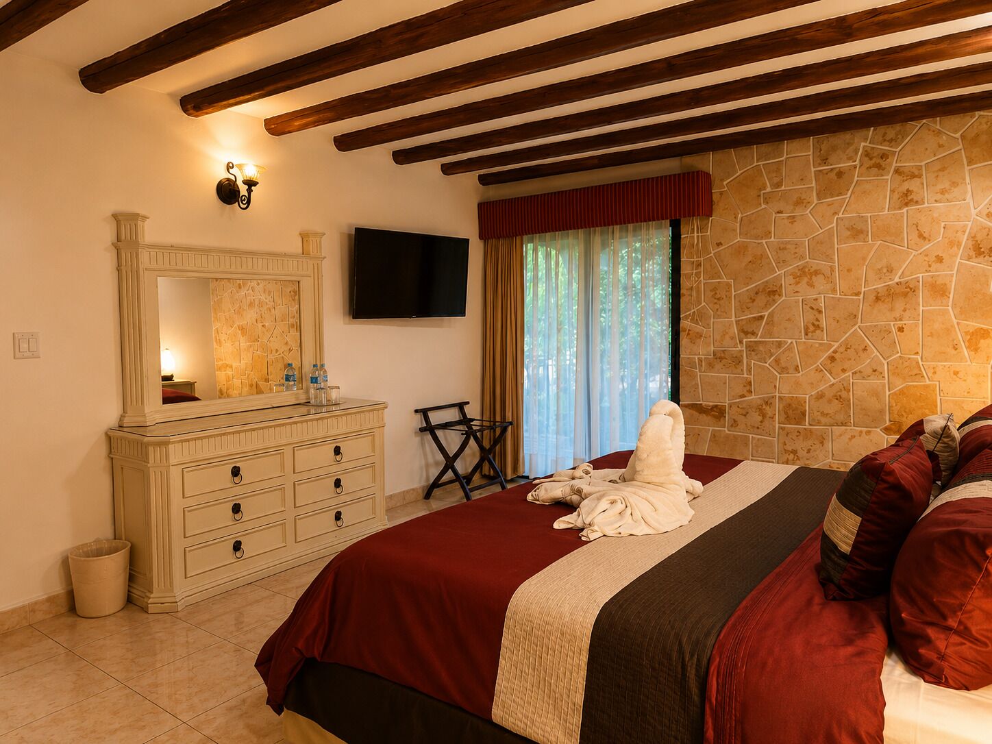 Quarto com parede de pedra, cama com roupa de cama vermelha, teto de madeira, TV na parede e cômoda com espelho.