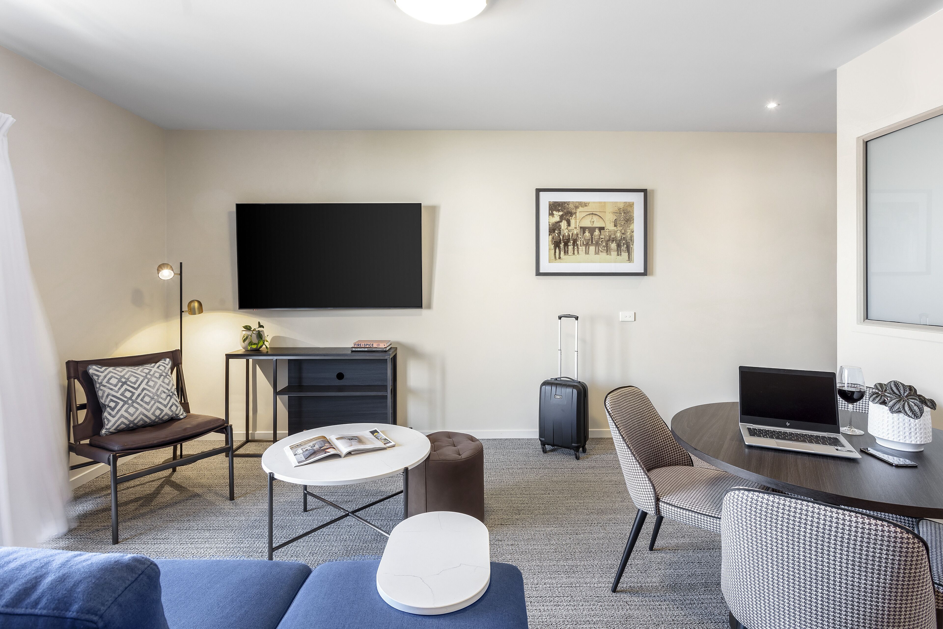 Quarto de hotel moderno com televisão de tela plana, uma mesa de jantar com um laptop, um sofá, uma cadeira e um pequeno banquinho.