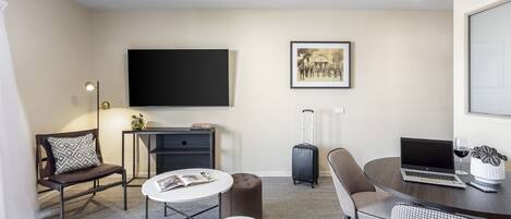 Quarto de hotel moderno com televisão de tela plana, uma mesa de jantar com um laptop, um sofá, uma cadeira e um pequeno banquinho.