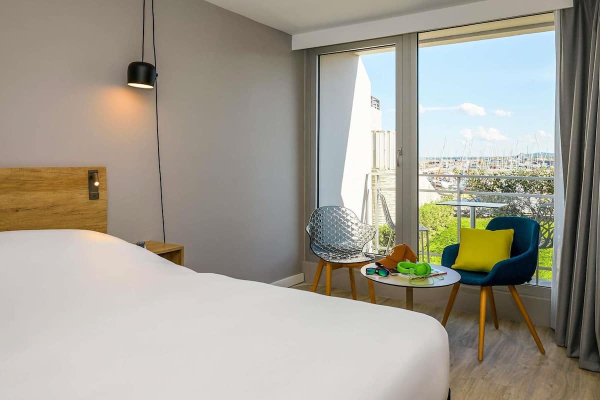 Quarto de hotel com cama, uma pequena mesa com cadeiras, varanda com vista para a marina e uma janela grande.