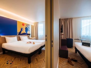 Quarto de hotel com duas camas, piso de madeira em padrão espinha de peixe, um quadro na parede, uma televisão e uma pequena mesa com uma garrafa de água.