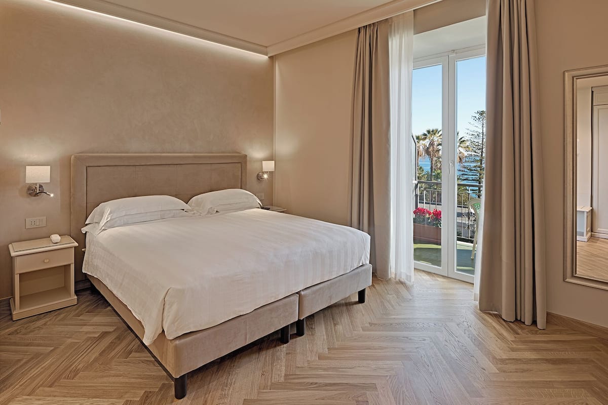 Quarto de hotel com uma cama grande, uma mesa de cabeceira, uma luminária e vista para o exterior através de uma porta de correr.