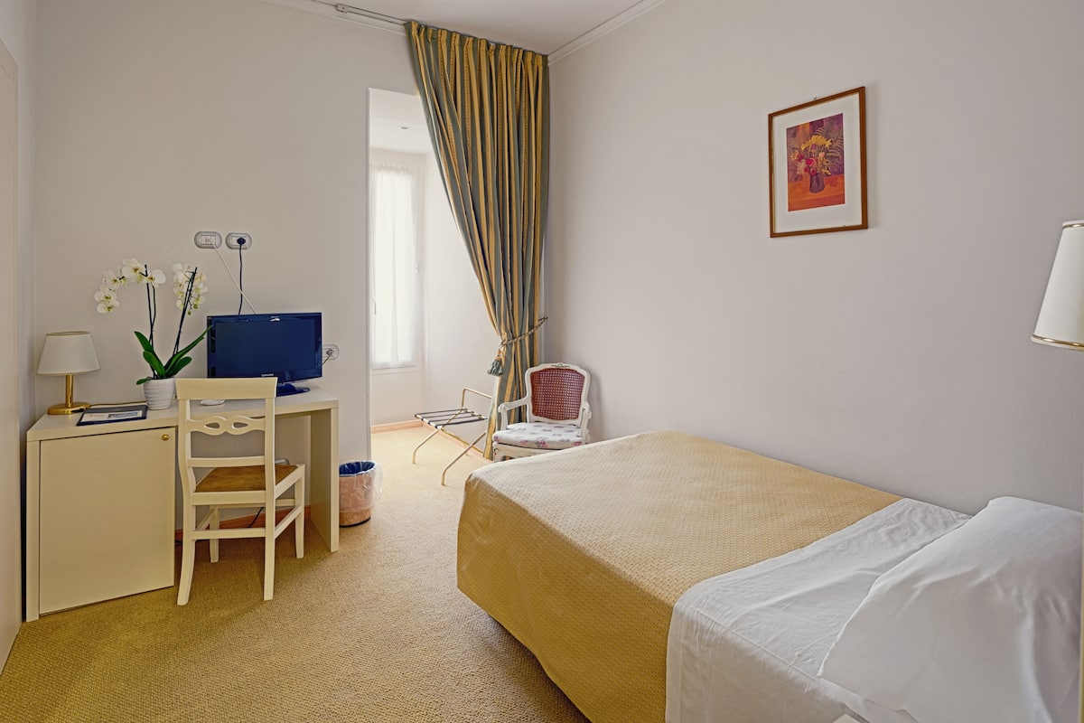 Quarto de hotel com cama, escrivaninha, cadeira, computador e um quadro na parede.