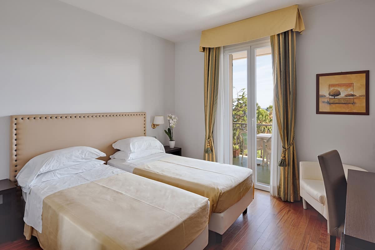 Quarto de hotel com duas camas, uma mesinha com abajur, uma cadeira e uma varanda com cortinas.