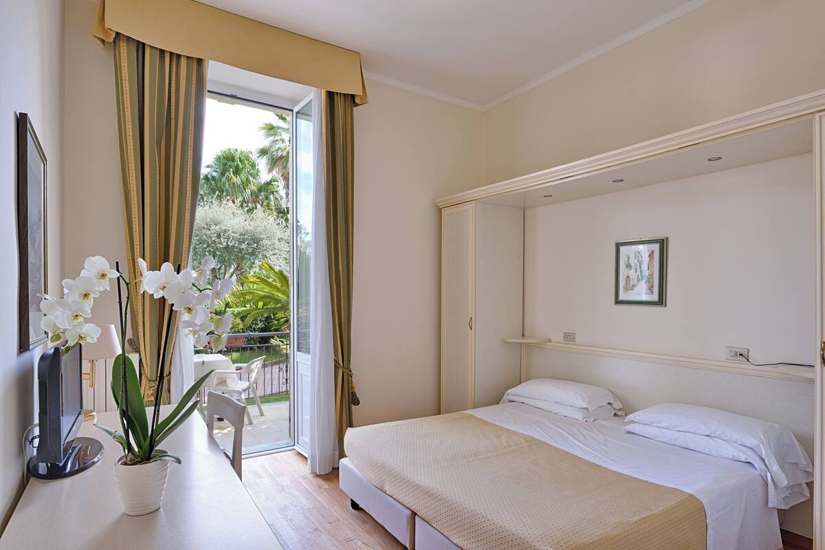 Quarto de hotel com cama, escrivaninha com computador, planta em vaso e vista para o exterior.