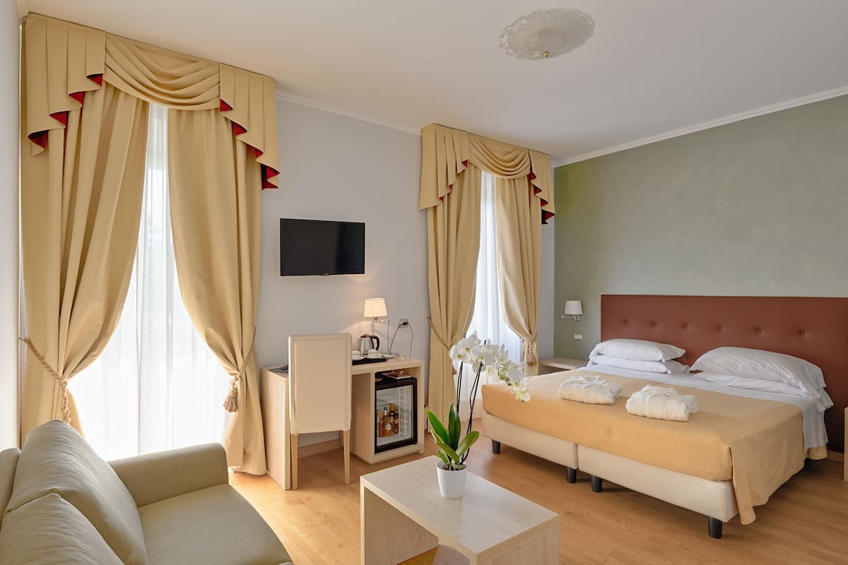 Quarto de hotel com cama, sofá, uma mesinha com uma planta, televisão e minibar.