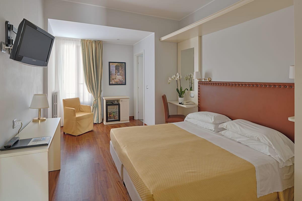 Quarto de hotel com cama, escrivaninha com luminária, cadeira, televisão na parede e pequena mesa com luminária.