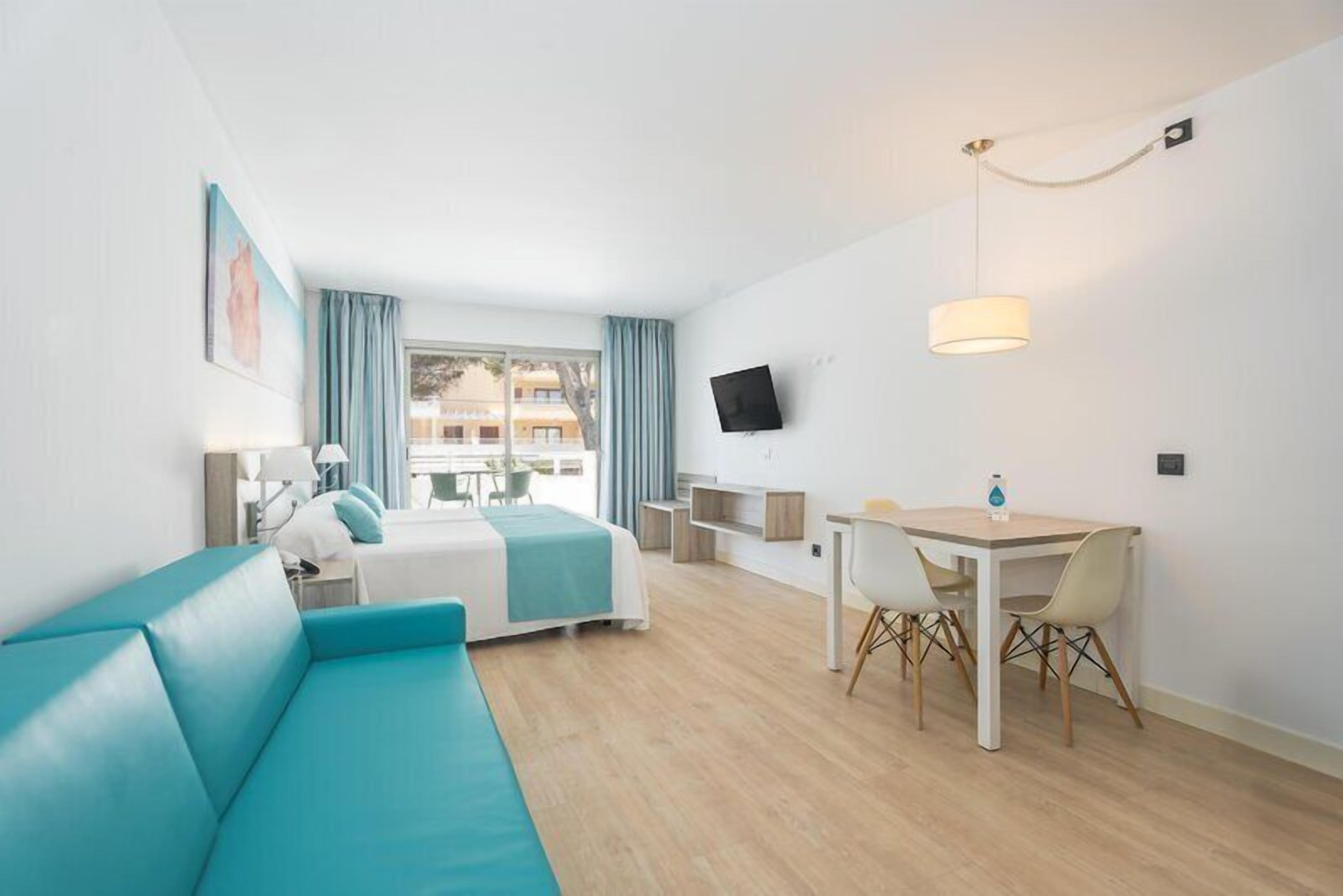 Quarto de hotel moderno com cama, sofá azul, mesa de jantar com cadeiras, televisão e janela com cortinas.