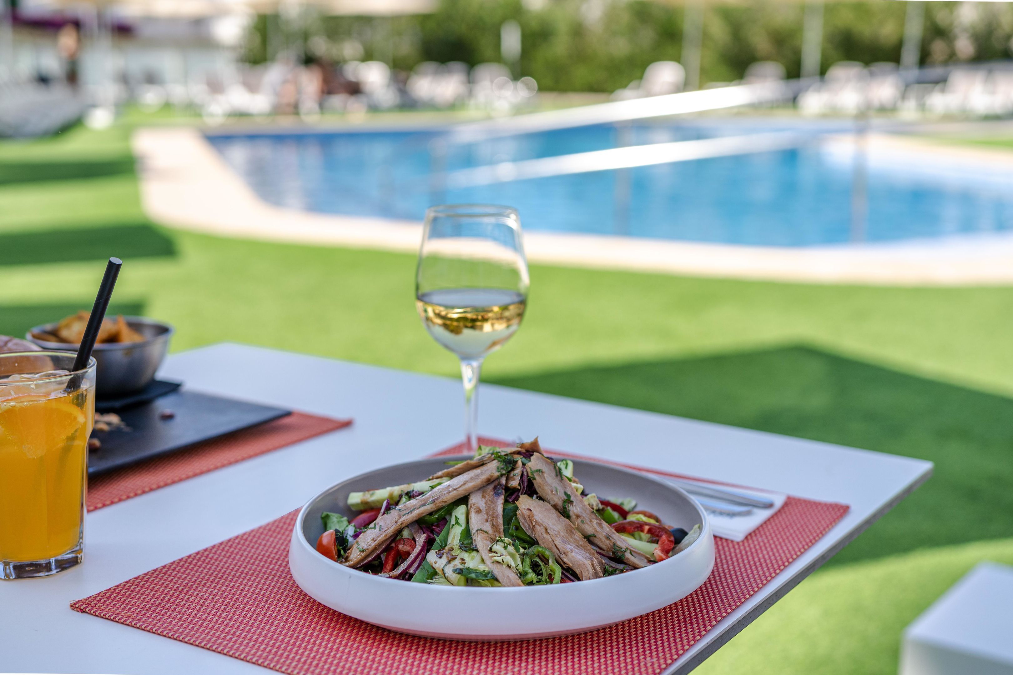 Uma mesa de jantar com uma salada, um copo de vinho branco e um copo de suco de laranja, montada ao ar livre com uma piscina ao fundo.