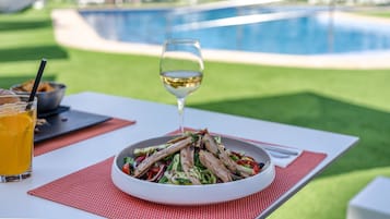 Uma mesa de jantar com uma salada, um copo de vinho branco e um copo de suco de laranja, montada ao ar livre com uma piscina ao fundo.