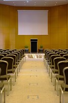 Sala de conferências com fileiras de cadeiras voltadas para um palco com uma tela grande.