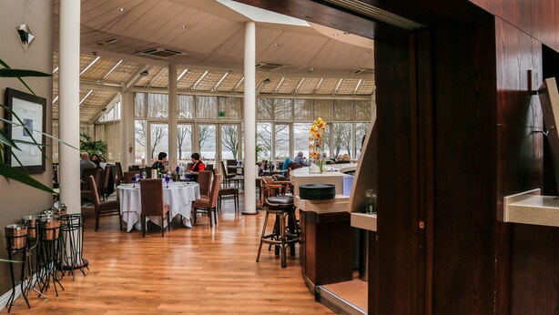 Um restaurante amplo, com pé direito alto, janelas grandes e piso de madeira.