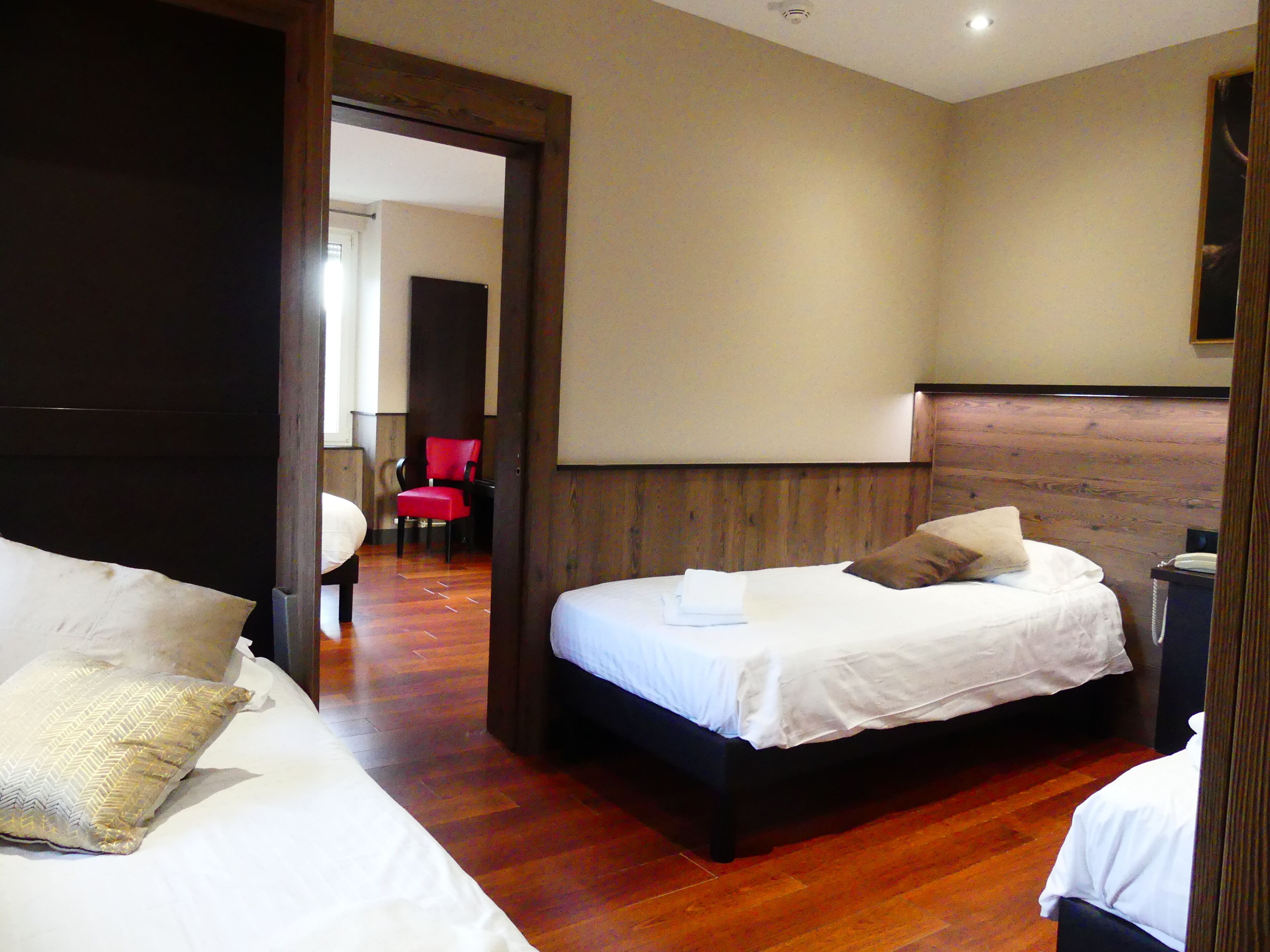 Quarto de hotel com duas camas, cabeceira de madeira, uma cadeira vermelha e piso de madeira.