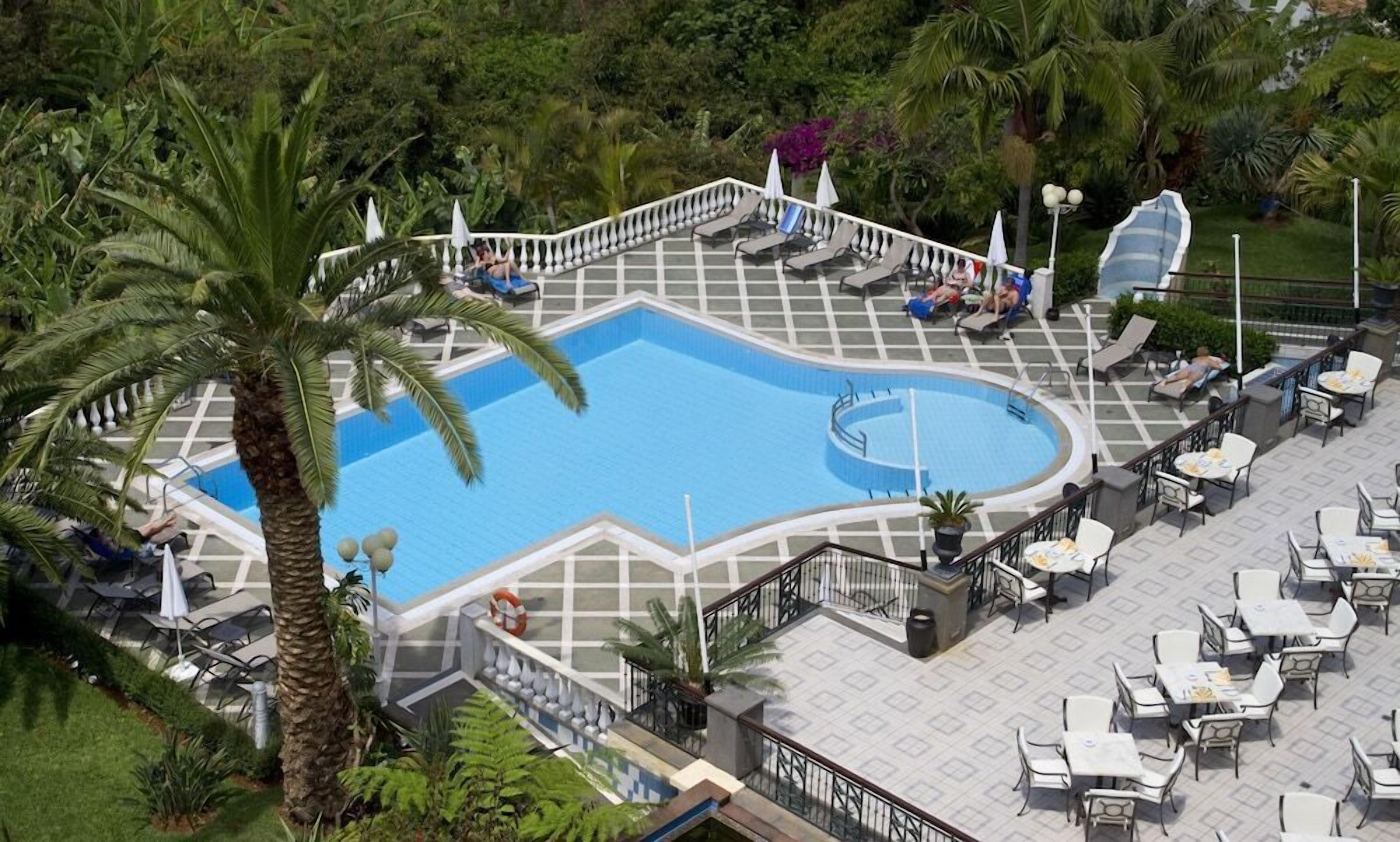 Uma grande piscina ao ar livre cercada por espreguiçadeiras e guarda-sóis, com palmeiras e um deck revestido de azulejos.