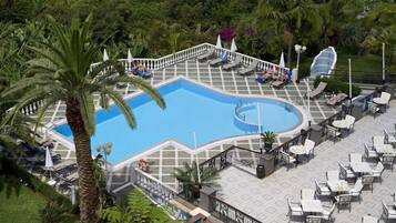 Uma grande piscina ao ar livre cercada por espreguiçadeiras e guarda-sóis, com palmeiras e um deck revestido de azulejos.
