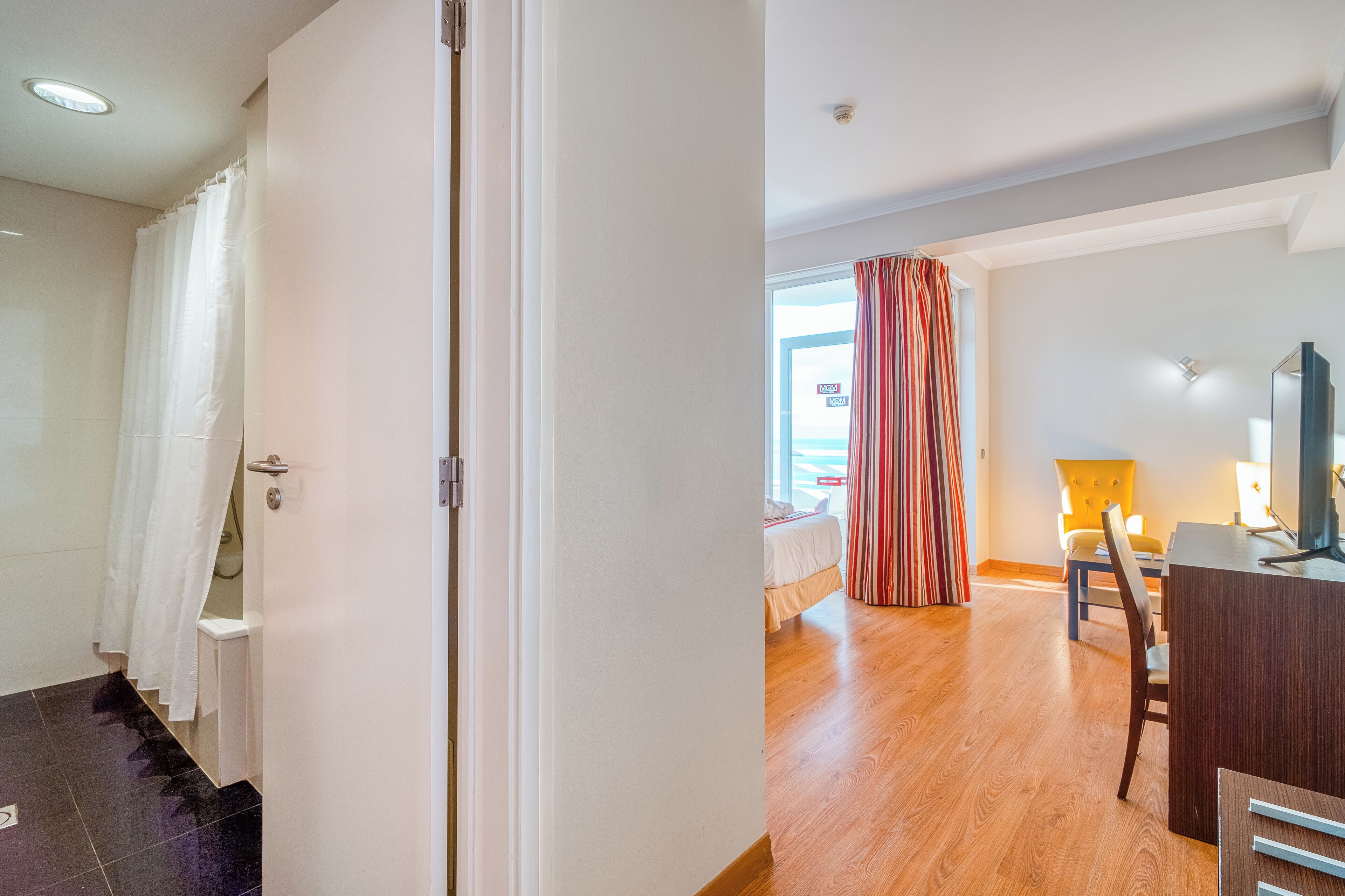 Quarto de hotel com piso de madeira, cama com roupa de cama branca, escrivaninha com cadeira e banheiro com vaso sanitário e chuveiro.