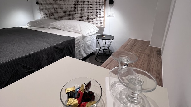 Quarto de hotel moderno com uma cama bem arrumada, uma pequena mesa de cabeceira com uma tigela de vidro cheia de chocolates e uma mesinha de madeira com um decantador de vidro.