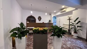 Uma área de recepção com piso de mármore, plantas em vasos, balcão de atendimento e uma estante de livros.