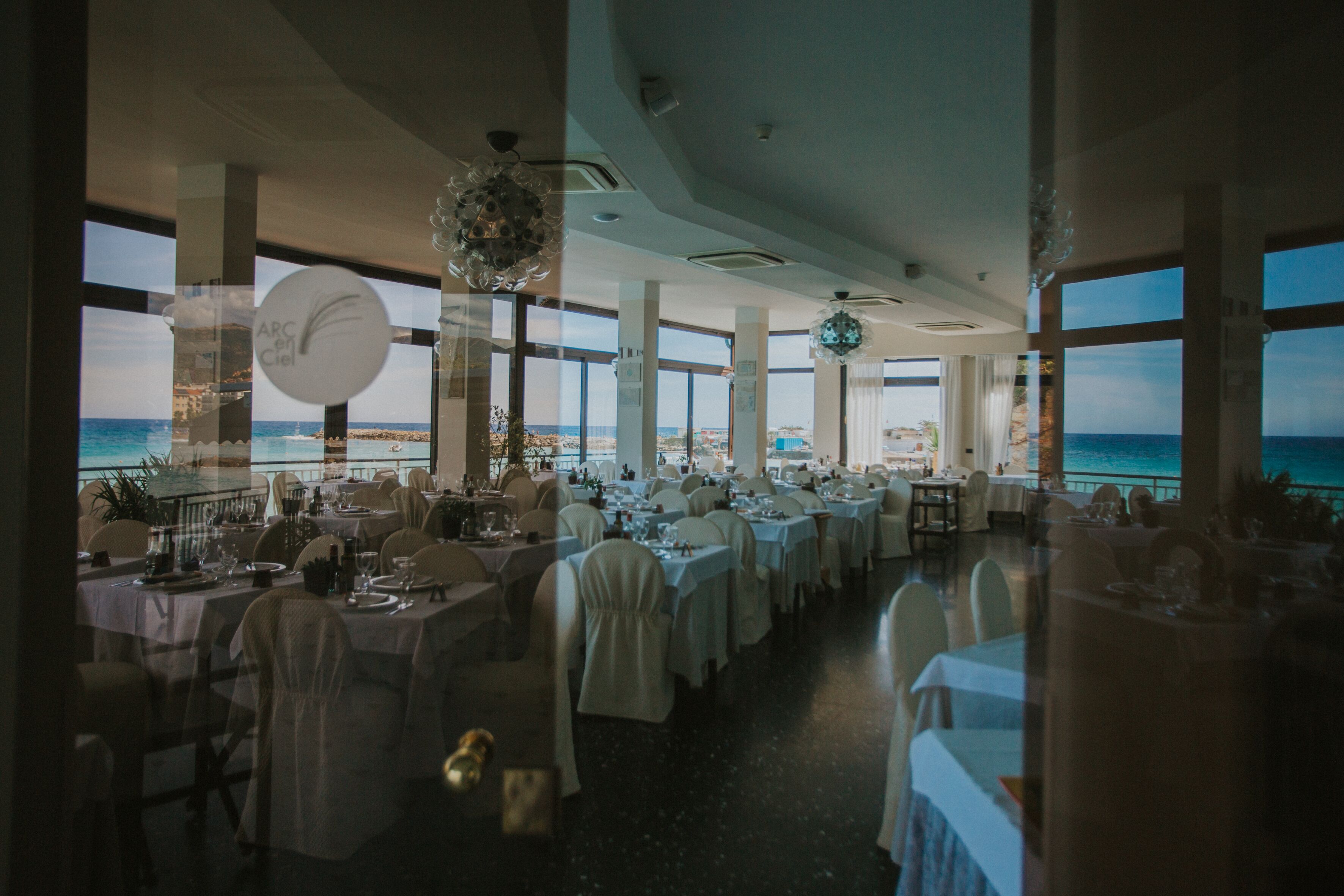 Restaurante com mesas preparadas para refeições, amplas janelas com vista para o mar e design de interiores moderno.