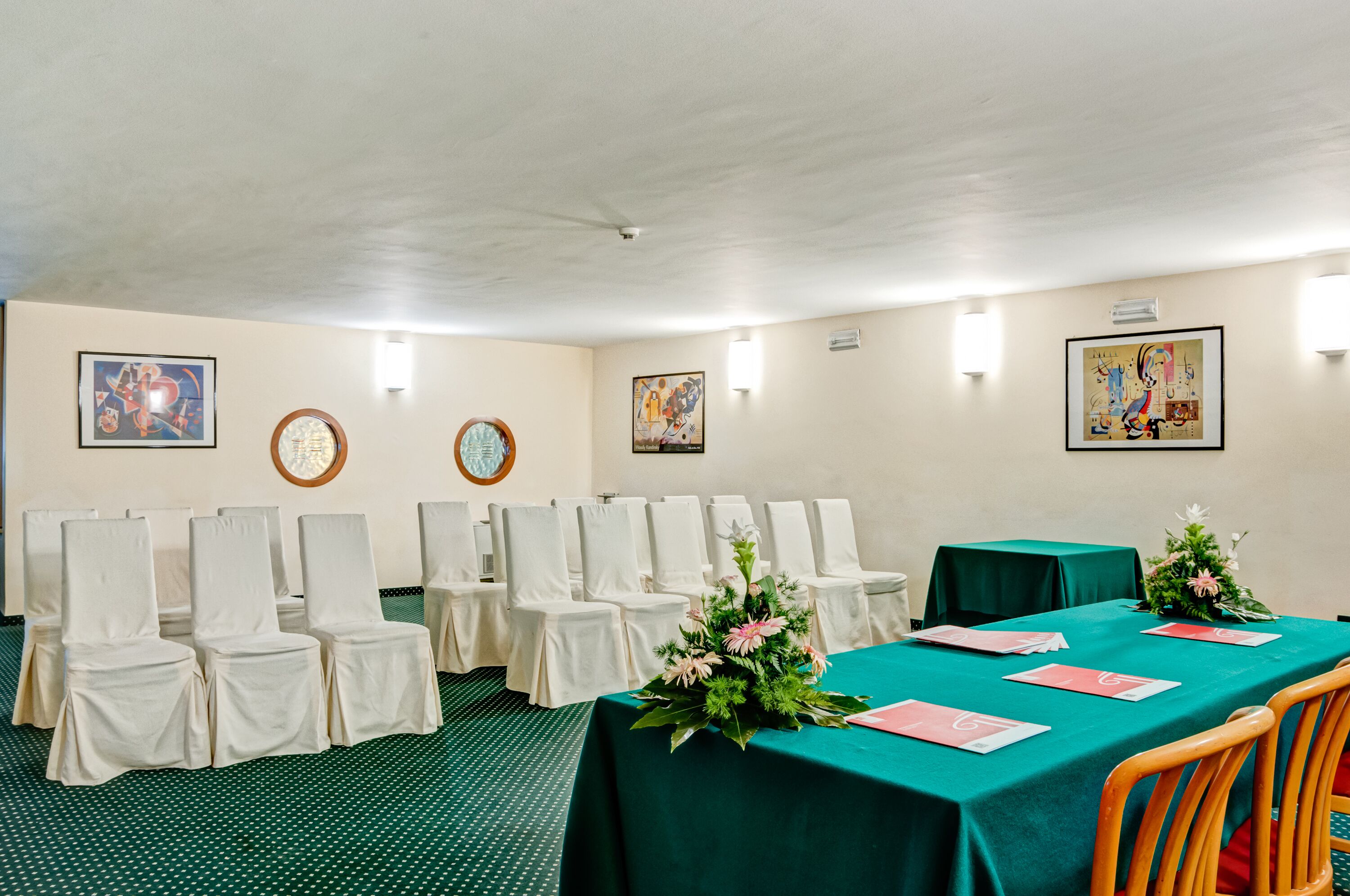 Uma sala de conferências com fileiras de cadeiras brancas, uma mesa verde com arranjos florais e obras de arte nas paredes.
