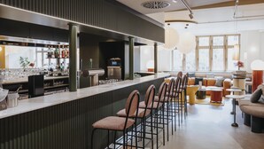 Uma área de bar moderna com um balcão longo, banquetas e um espaço de estar com sofás e mesas.