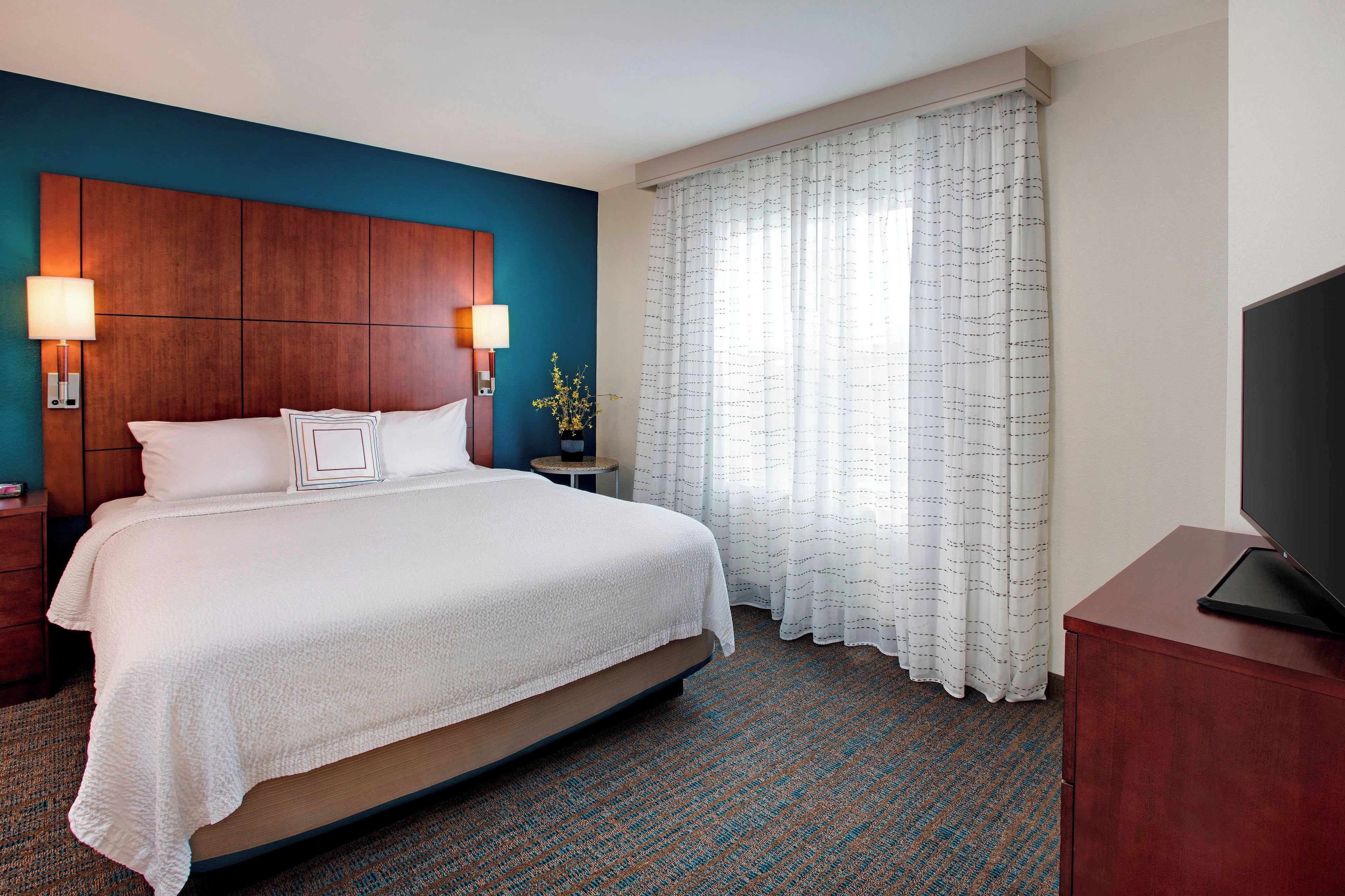 Quarto de hotel com uma cama grande, duas luminárias de cabeceira, uma TV de tela plana, uma mesinha com um vaso de flores e uma janela com cortinas translúcidas.
