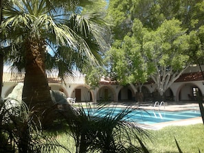 Uma área de piscina com uma piscina azul cristalina, rodeada por palmeiras e um edifício com portas em arco.