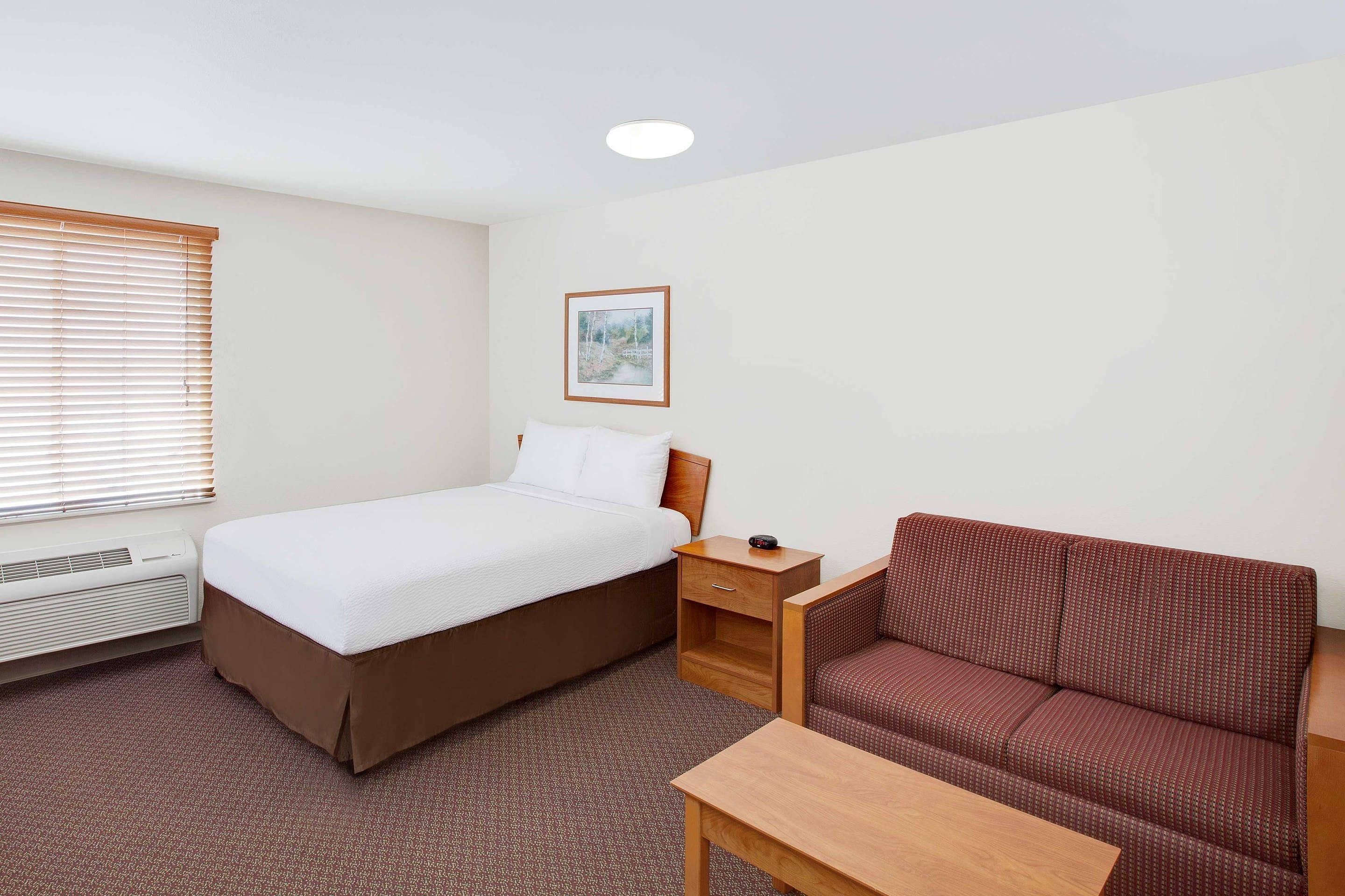 Quarto de hotel com cama, mesa de cabeceira, sofá vermelho estampado, pequena mesa de madeira e quadro na parede.