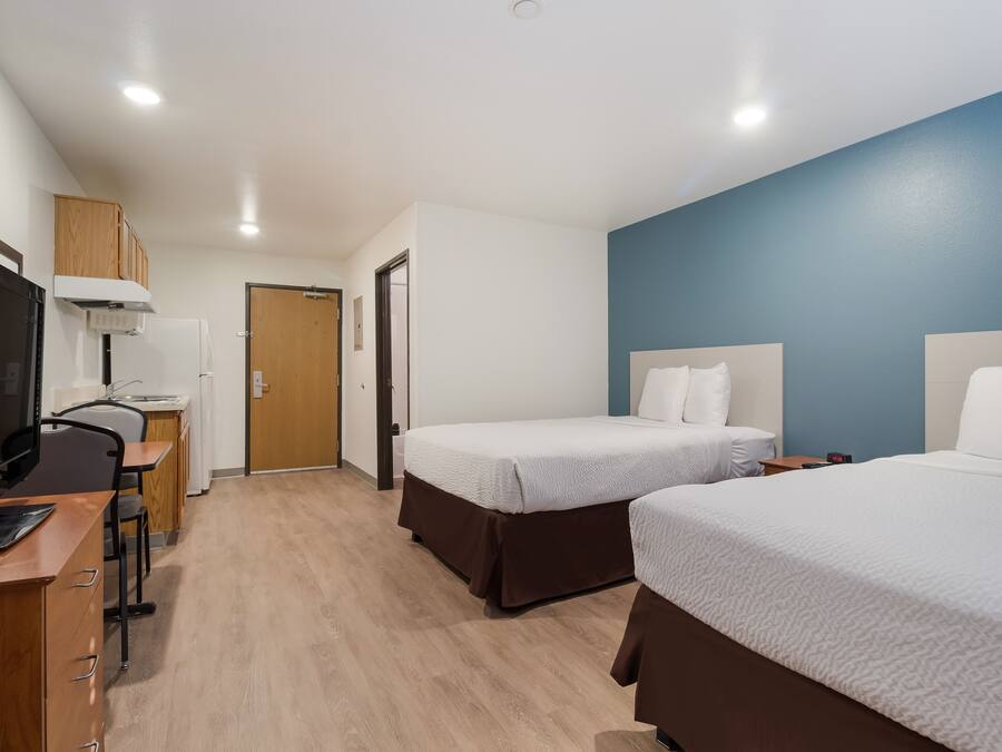Quarto de hotel com duas camas, uma escrivaninha com computador, televisão e cozinha compacta.