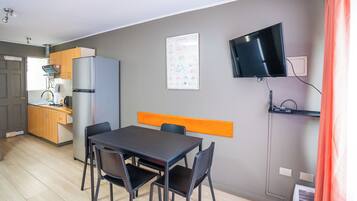 Cozinha compacta com geladeira, fogão e pia. Há uma área de refeições com mesa e cadeiras, e uma televisão fixada na parede.