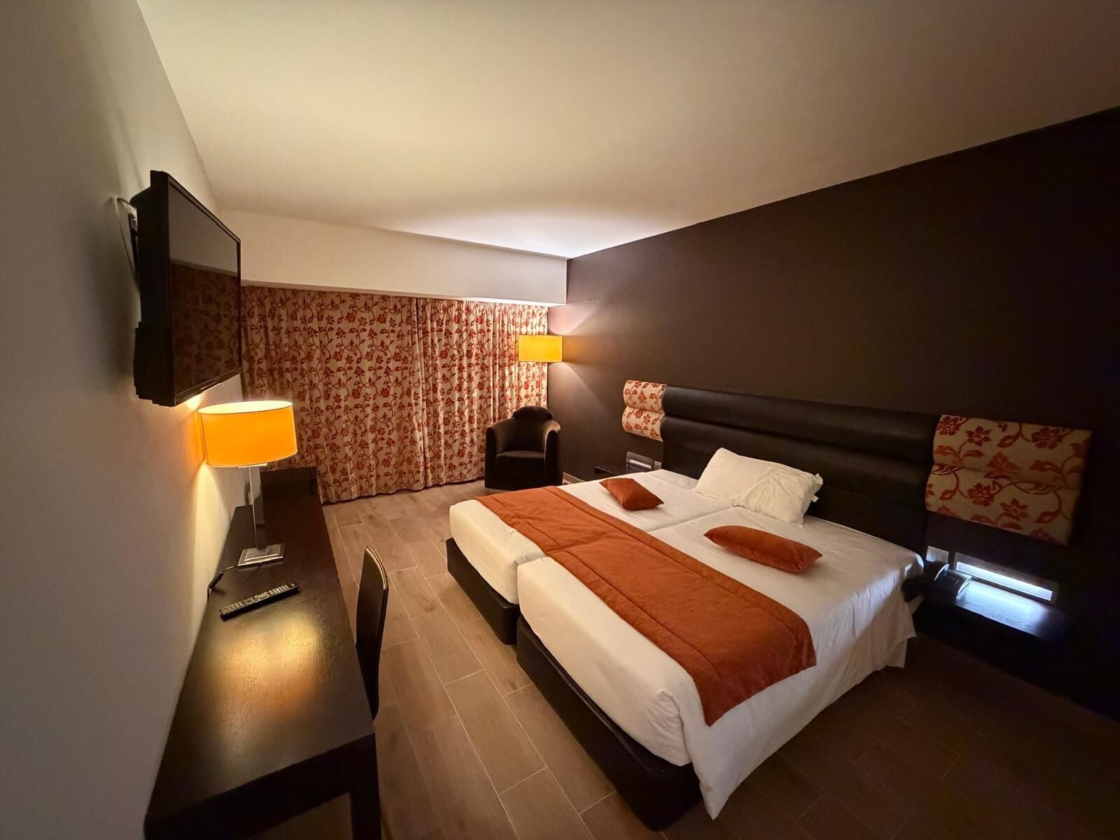 Quarto de hotel com uma cama grande, duas mesas de cabeceira, uma escrivaninha com cadeira, uma TV montada na parede e uma janela com cortinas.