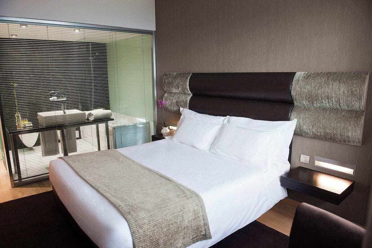 Quarto de hotel moderno com uma cama grande, mesa de cabeceira, persianas e vista para o exterior.
