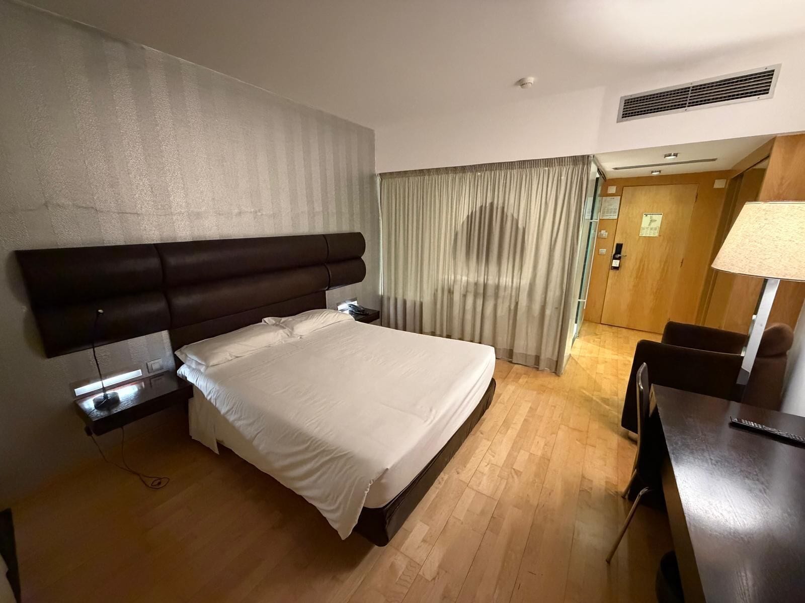 Quarto de hotel com uma cama grande, uma mesa de cabeceira de madeira, uma escrivaninha com luminária e uma porta que dá para outro espaço.