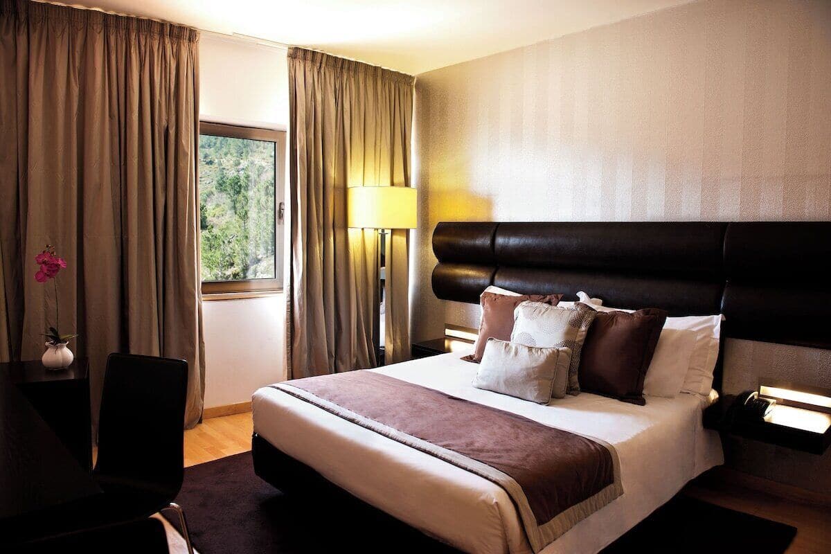 Quarto de hotel com uma cama grande, mesa de cabeceira, abajur, cadeira e janela com cortinas.