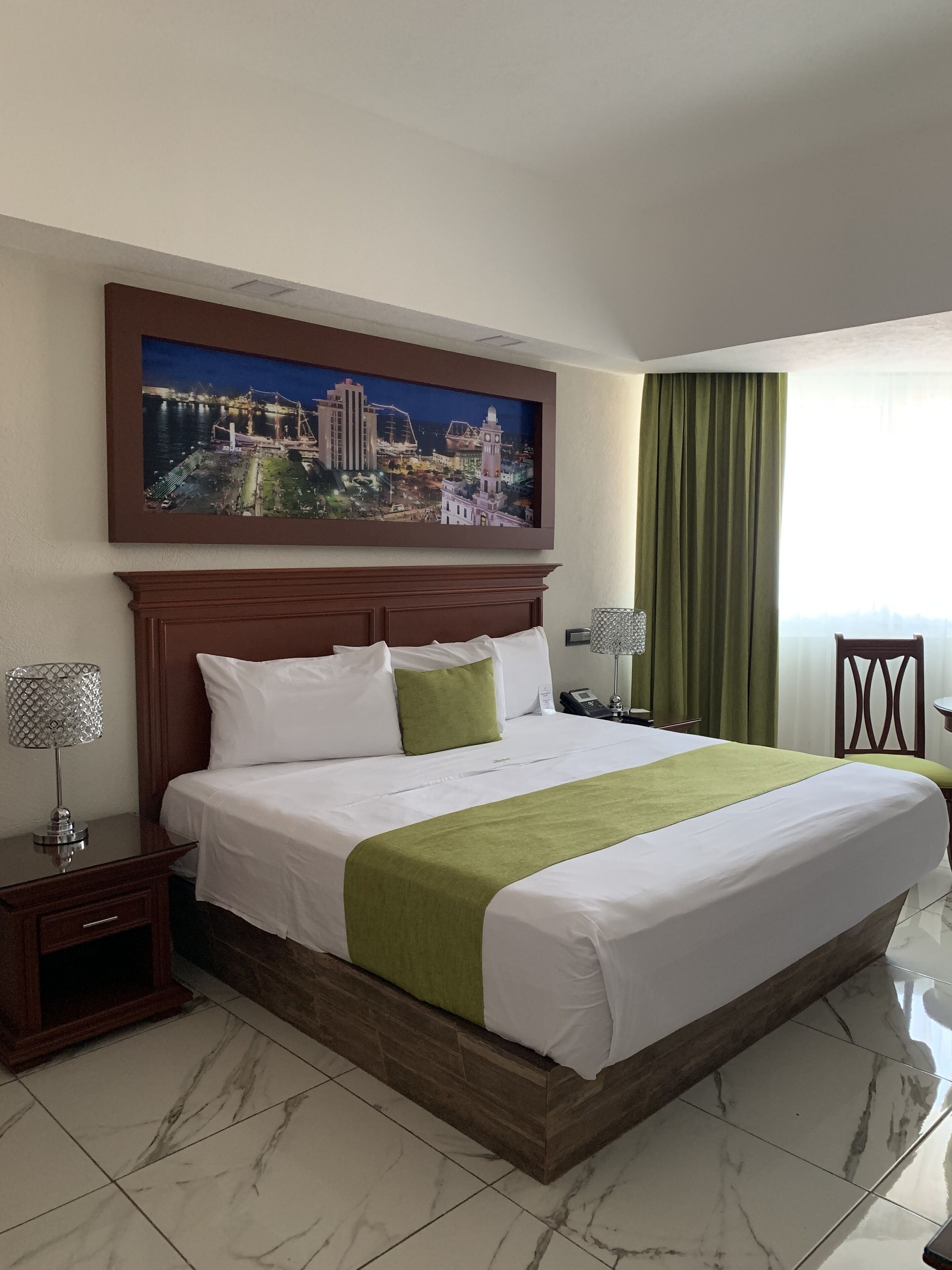 Quarto de hotel com uma cama grande, uma mesa de cabeceira, um abajur, um quadro com vista urbana e vista para o exterior.