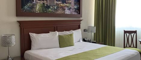 Quarto de hotel com uma cama grande, uma mesa de cabeceira, um abajur, um quadro com vista urbana e vista para o exterior.
