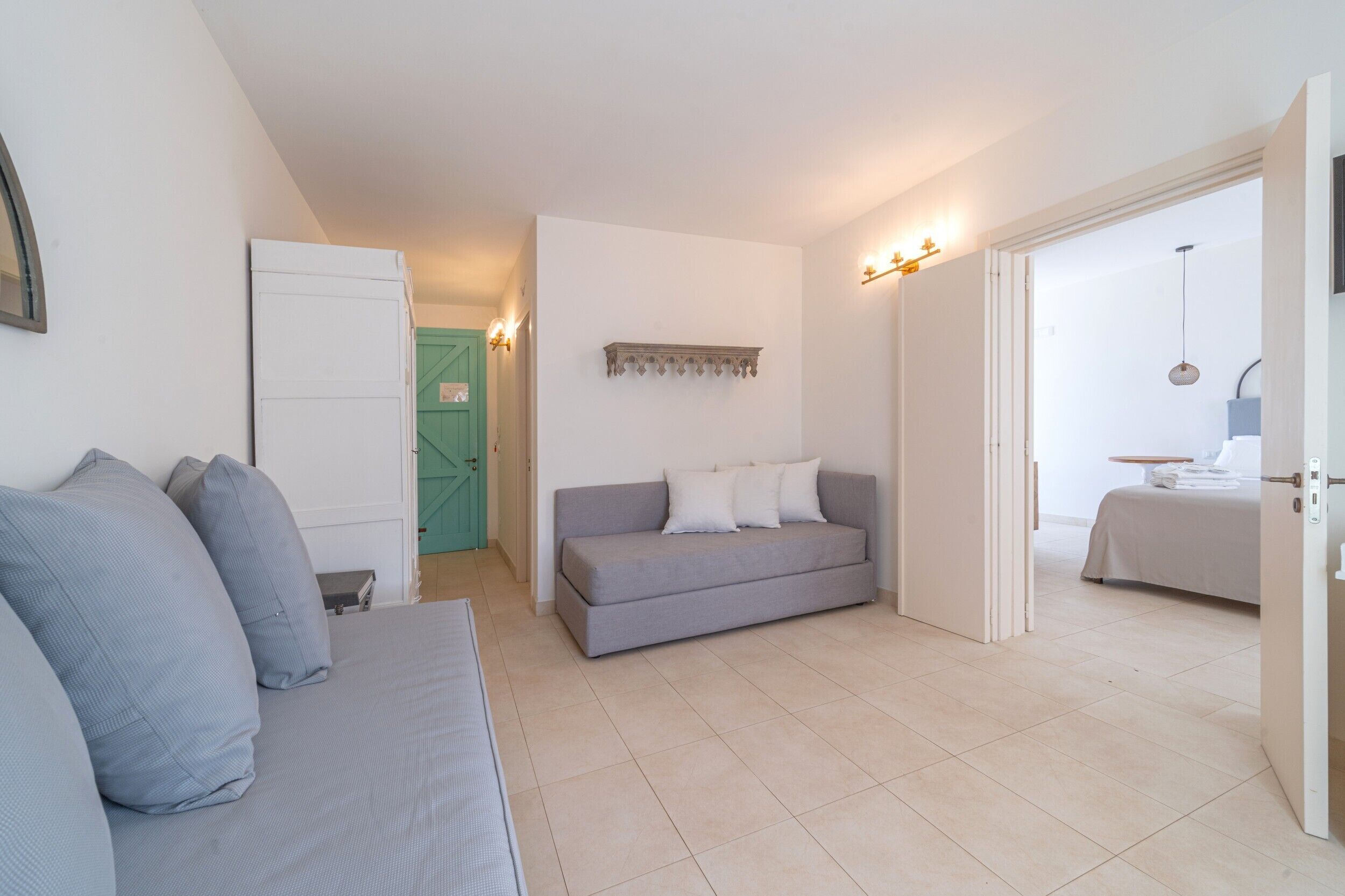 Uma sala de estar moderna com um sofá cinzento, uma porta branca e uma cama ao fundo.