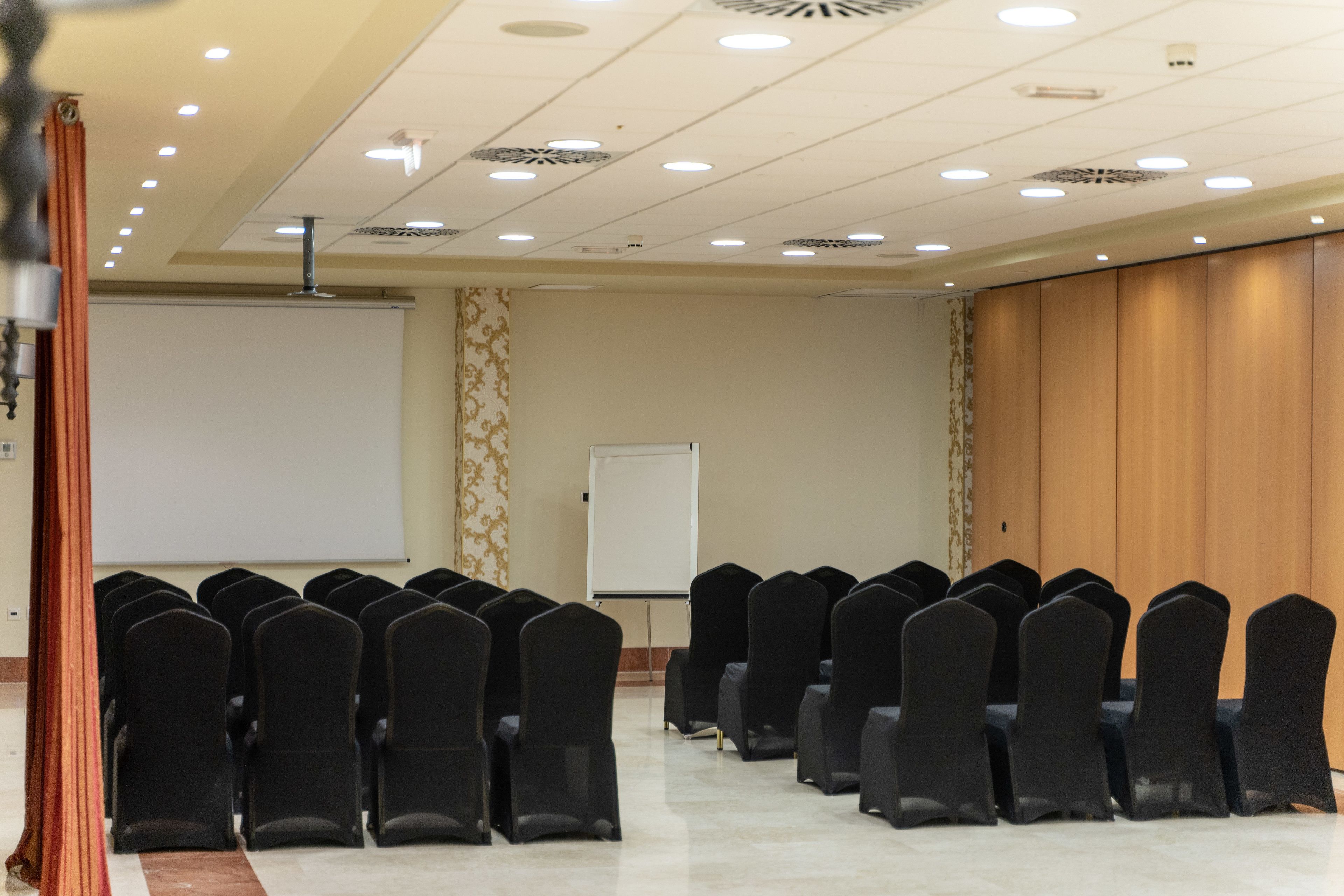 Uma sala de conferências com fileiras de cadeiras pretas, um quadro branco e uma tela de projeção.