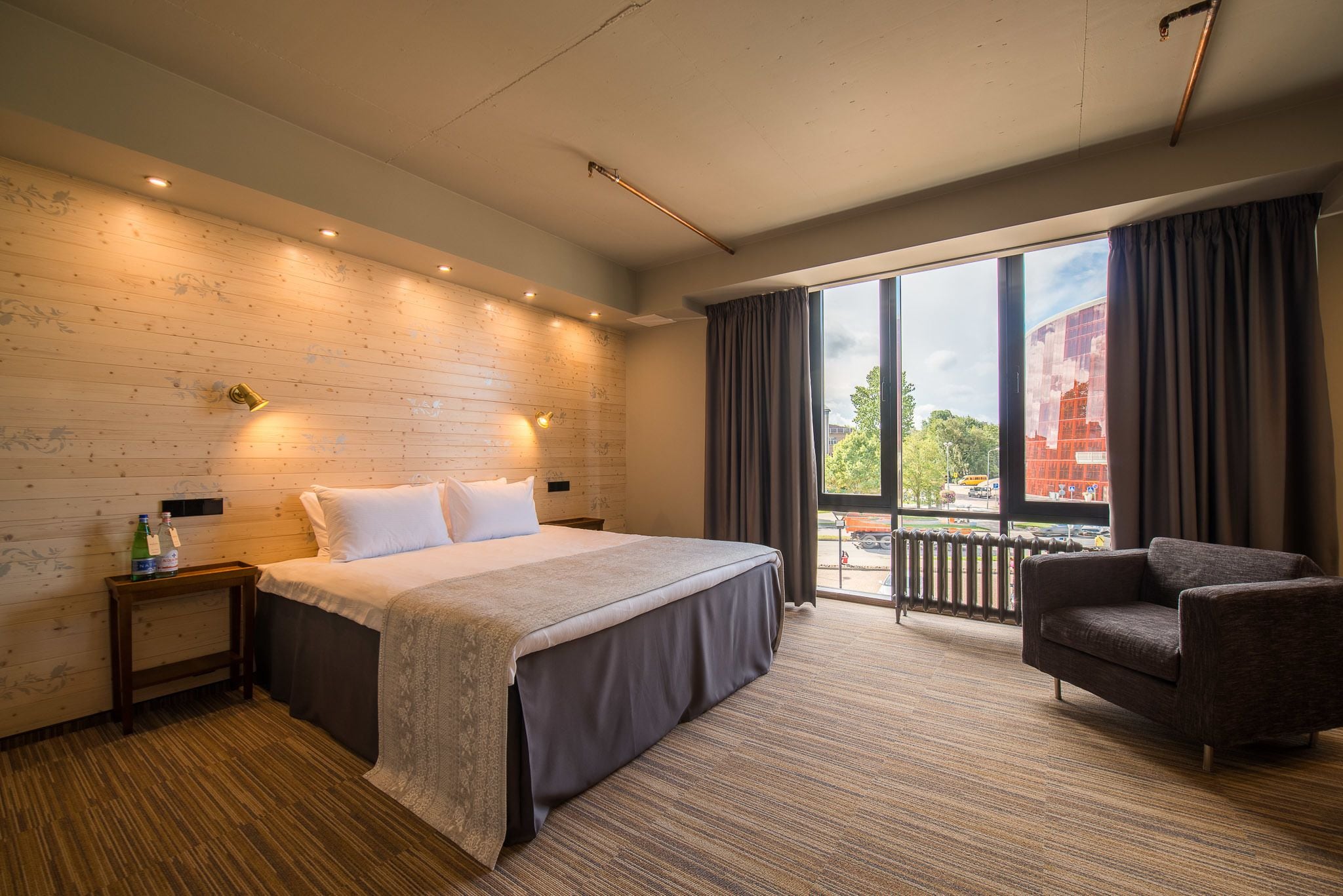 Quarto de hotel com uma cama grande, uma cadeira, uma mesinha de cabeceira e vista para o exterior.
