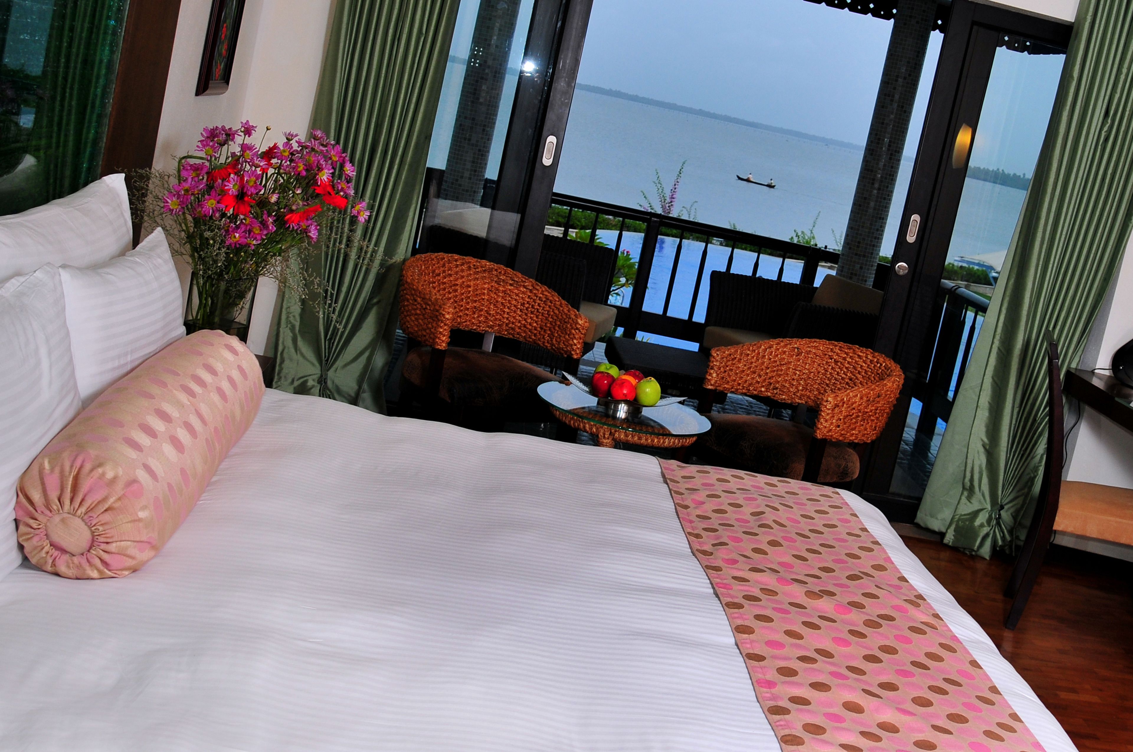 Quarto de hotel com uma cama, uma mesa pequena com frutas, duas cadeiras de vime e vista para o mar.