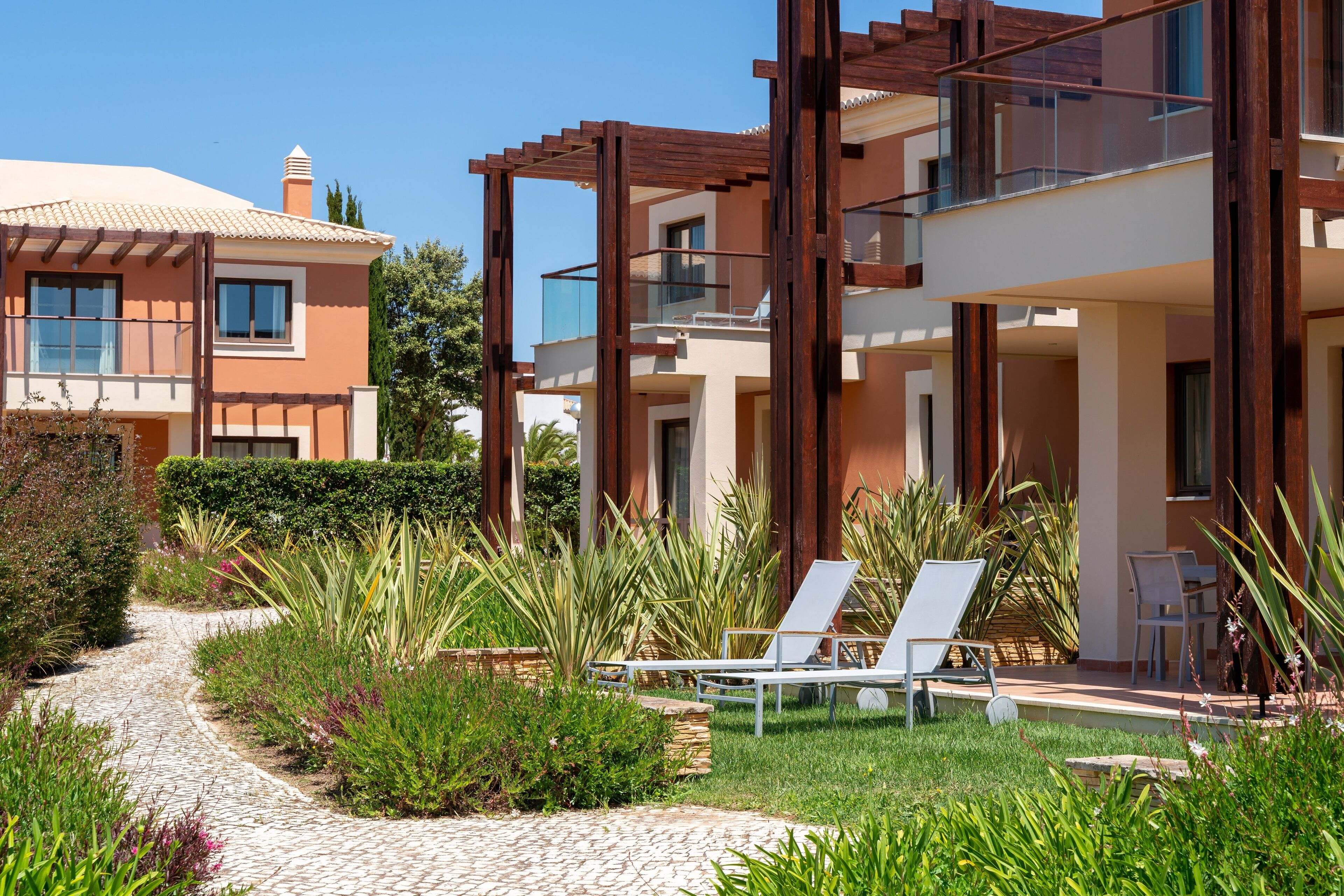 Um edifício moderno com caminho de pedra, vegetação exuberante e área de piscina com espreguiçadeiras.