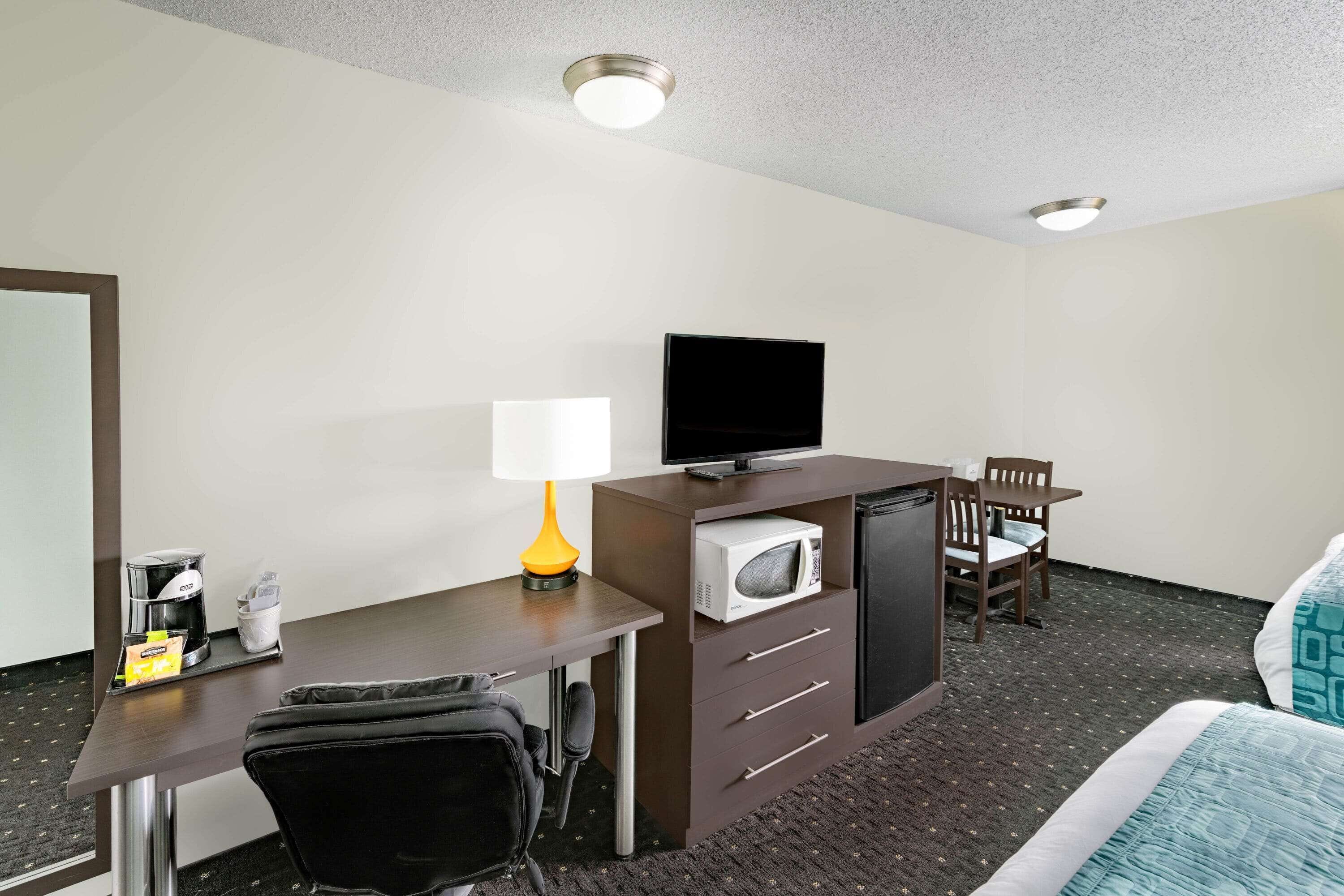 Quarto de hotel com uma cama, uma escrivaninha com abajur, micro-ondas, televisão e uma cadeira.