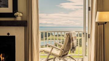 Um quarto aconchegante com lareira, uma cadeira de balanço com manta, e vista para um lago e colinas através de uma porta aberta.