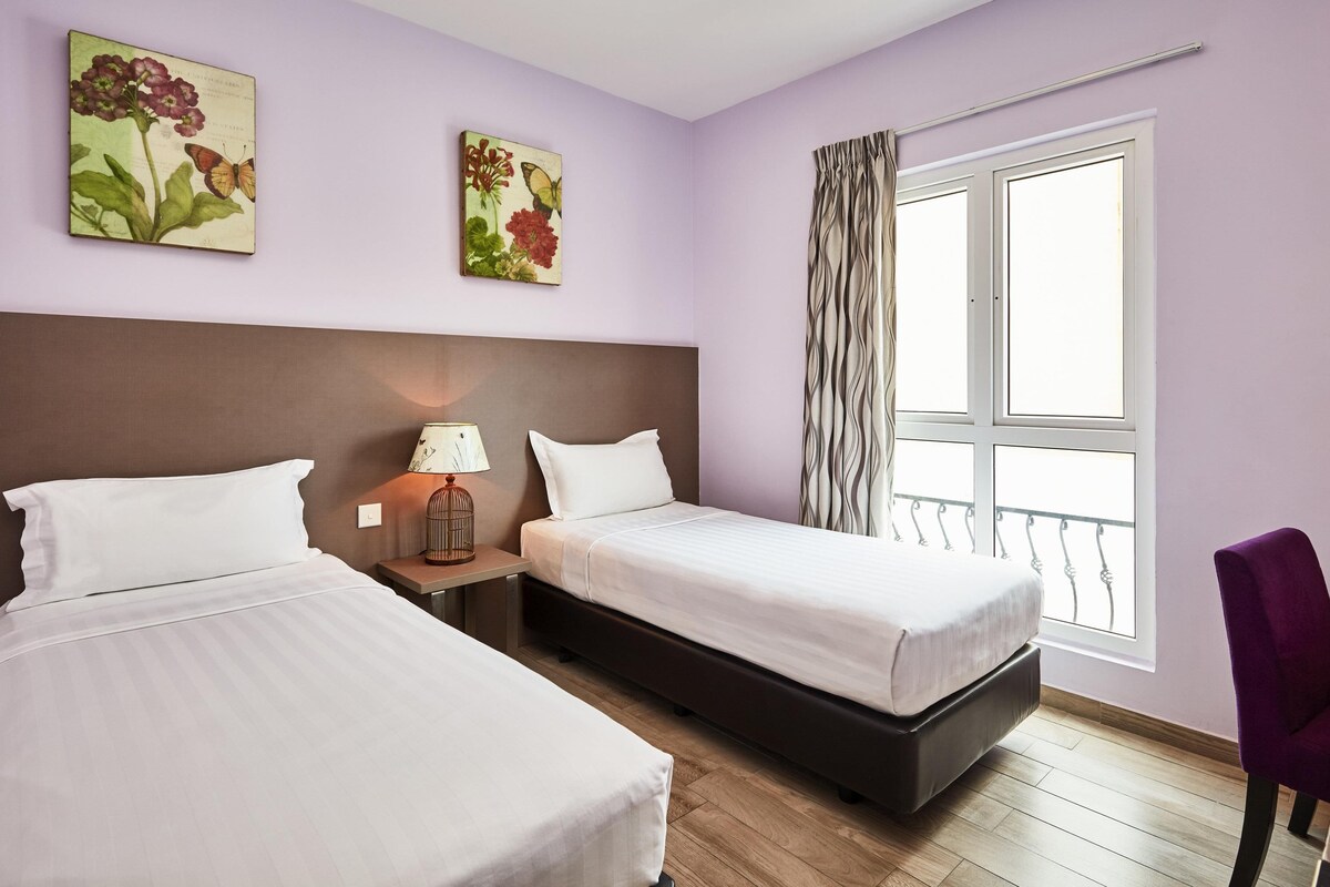 Quarto de hotel com duas camas, uma mesa de cabeceira com abajur e uma janela com cortinas.