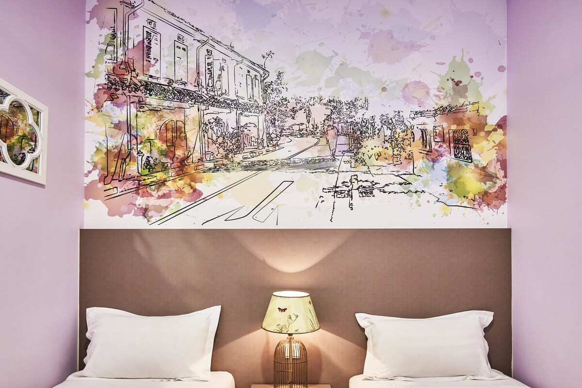 Quarto de hotel com uma cama, dois travesseiros, uma mesa de cabeceira com abajur e um mural na parede retratando uma cena de rua.