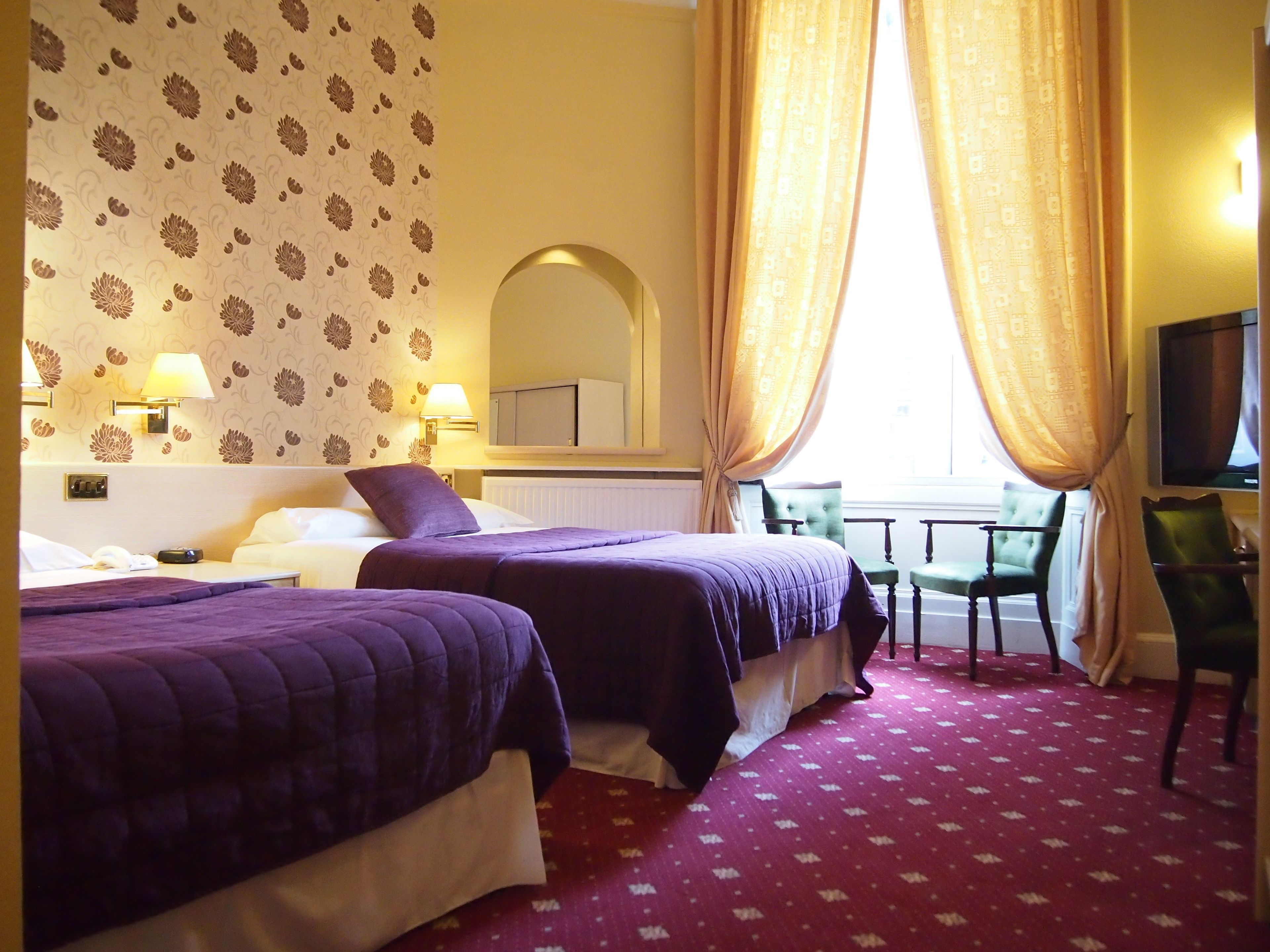 Quarto de hotel com duas camas, uma mesinha, duas cadeiras, televisão e janela com cortinas.